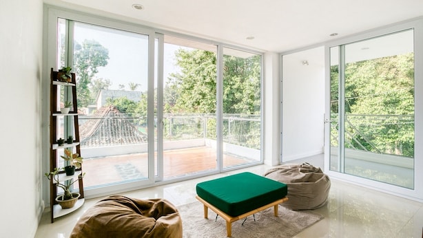 Quarto moderno com grandes portas de vidro deslizantes para uma varanda, um banco acolchoado verde, uma estante de madeira com plantas e um pufe bege.