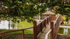 Uma ponte de madeira sobre águas calmas, cercada por vegetação exuberante e um edifício tradicional ao fundo.
