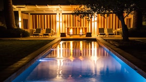 Piscina com espreguiçadeiras e área com lareira, rodeada por um pátio bem iluminado.