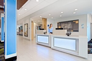 Uma área de recepção de hotel com design moderno, com balcão de recepção, um quiosque de check-in digital e uma placa fixada na parede indicando horários de entrada e saída.