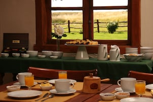 Mesa de café da manhã posta com diversos pratos, xícaras e um jarro, com vista para um campo gramado através de uma janela.