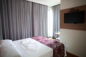 Quarto de hotel com cama, televisão fixada na parede e escrivaninha com cadeira.