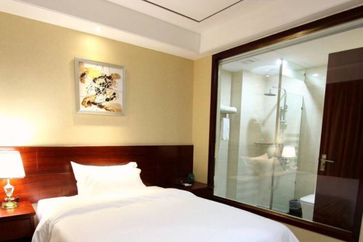 Quarto de hotel com uma cama grande, uma mesinha de cabeceira com abajur, um quadro na parede e um banheiro com divisória de vidro.