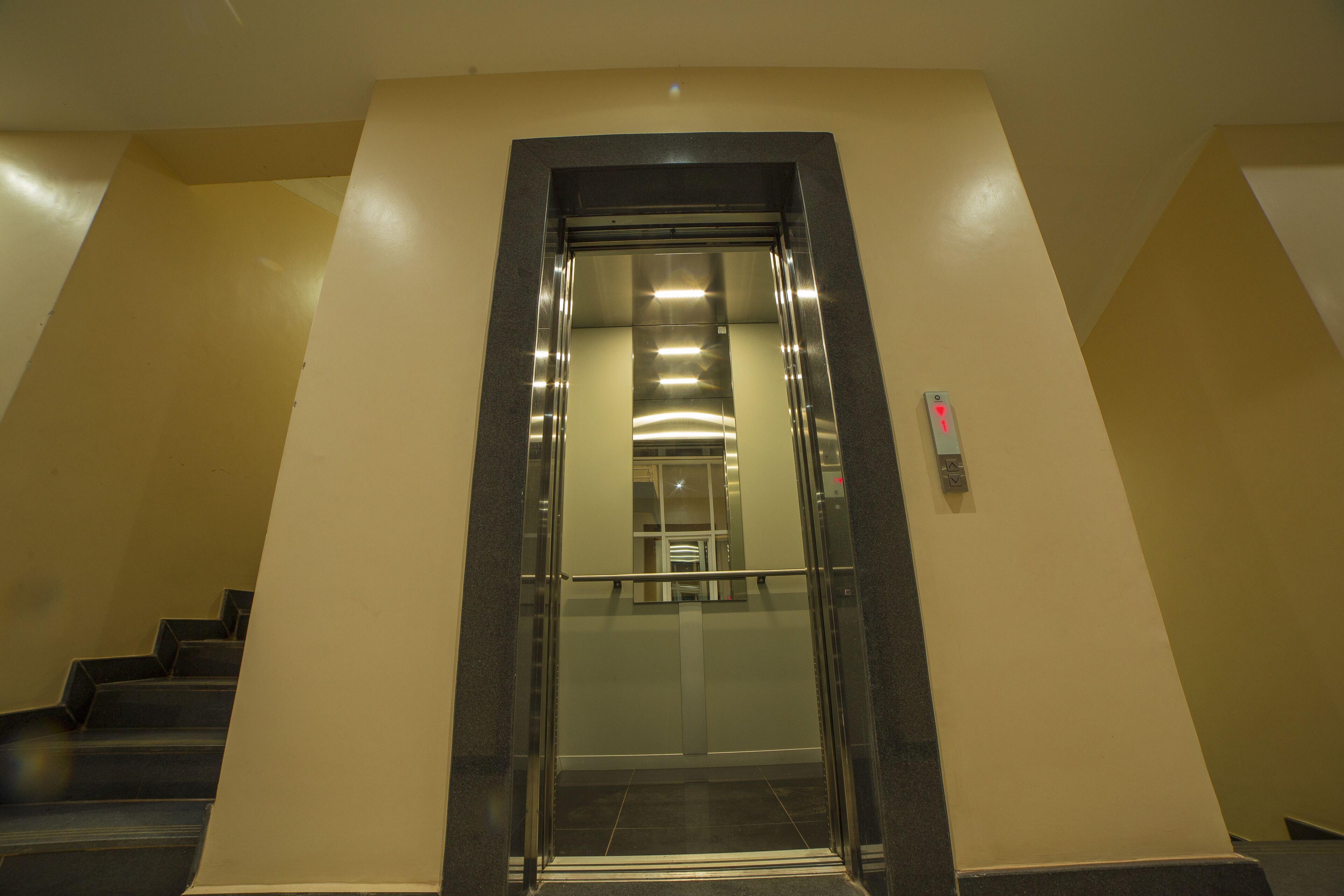 Entrada de elevador com porta de vidro e corrimão, dando acesso a escadaria com degraus escuros.