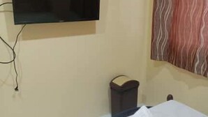 Quarto de hotel com cama, televisão fixada na parede, lixeira e cortina estampada.