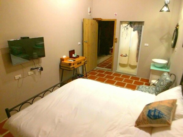 Quarto de hotel com cama, televisão fixada na parede, mesinha com abajur e banheiro com pia e espelho.