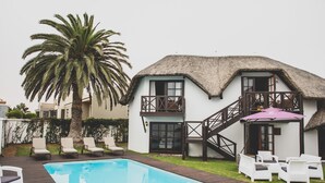 Área da piscina com um edifício de telhado de palha, cadeiras de descanso e uma palmeira.
