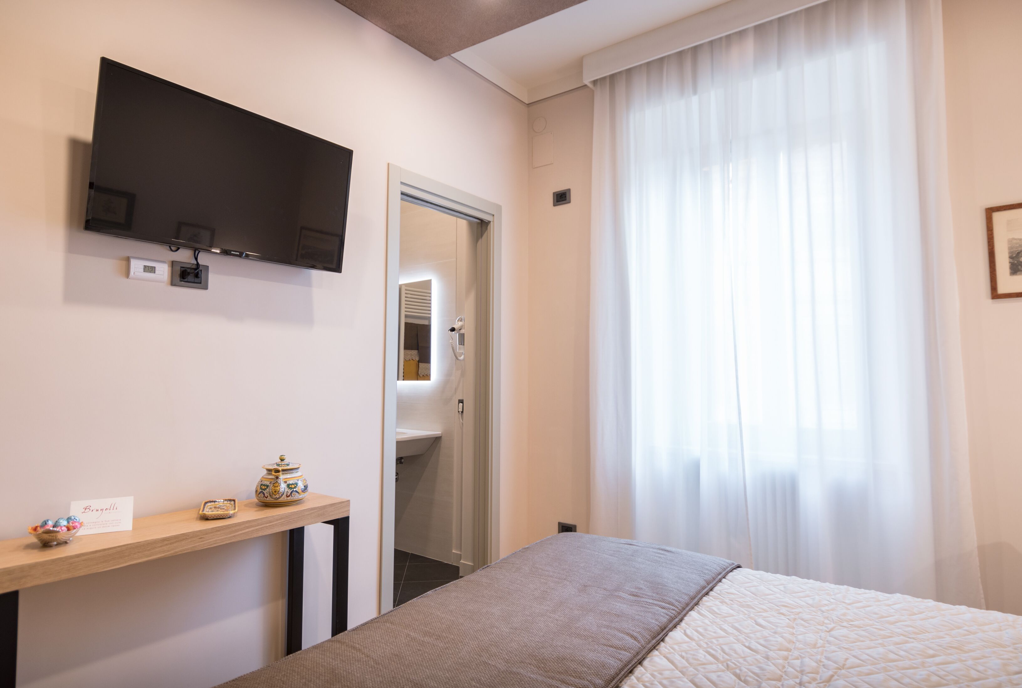 Quarto de hotel com cama, televisão na parede, uma mesinha com objetos decorativos e um banheiro visível através de uma porta aberta.