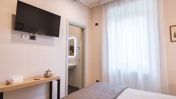 Quarto de hotel com cama, televisão na parede, uma mesinha com objetos decorativos e um banheiro visível através de uma porta aberta.