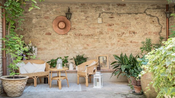Paredão rústico de pedra com um chapéu, móveis de vime e plantas em vasos numa área de estar ao ar livre acolhedora.