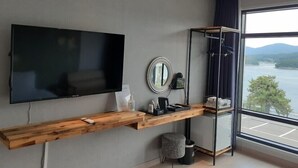 Quarto de hotel moderno com uma televisão grande fixada na parede, uma prateleira de madeira com produtos de higiene pessoal, um espelho e vista para um lago através de uma janela ampla.