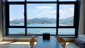 Quarto com uma grande janela que oferece vista para um lago e montanhas. Há uma mesa com cadeiras e um pequeno objeto preto sobre ela.
