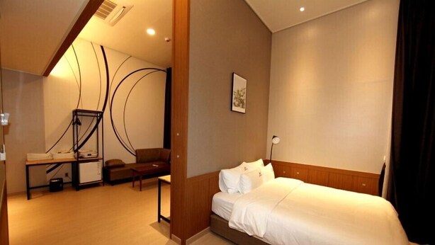 Quarto de hotel com cama, escrivaninha, cadeira e prateleira fixa na parede.