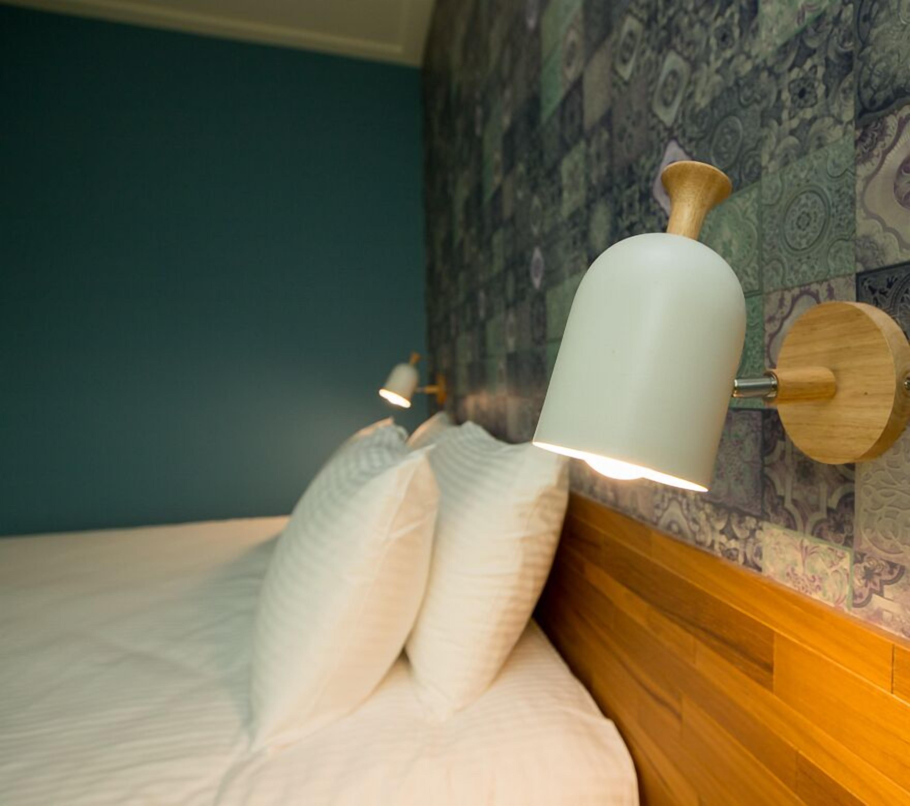 Uma cama bem arrumada com lençóis e almofadas brancos, cabeceira de madeira e luminária fixada na parede com cúpula branca.