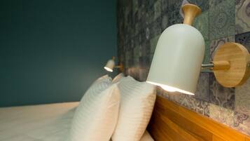Uma cama bem arrumada com lençóis e almofadas brancos, cabeceira de madeira e luminária fixada na parede com cúpula branca.