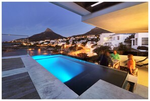 Área de piscina com vista para uma cidade costeira ao entardecer, apresentando arquitetura moderna e uma montanha ao fundo.