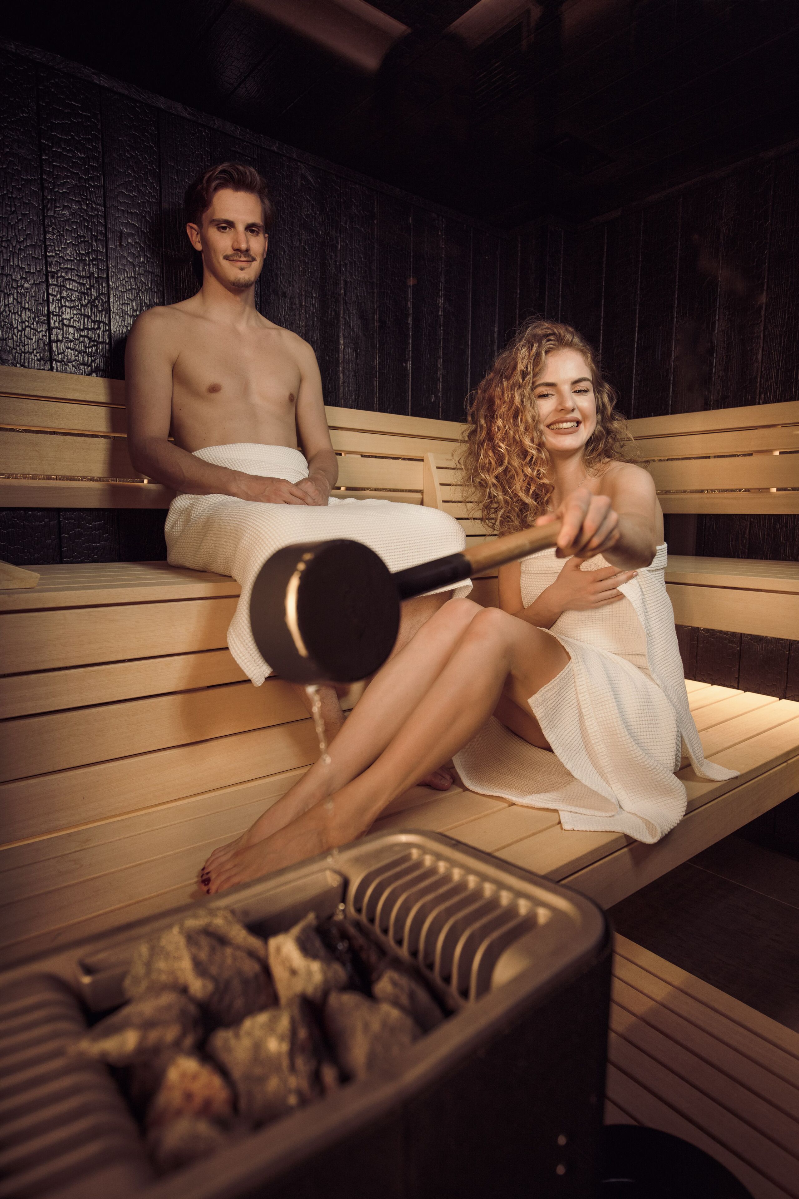Duas pessoas sentadas em bancos de madeira em um salão de sauna com parede de pedra e um recipiente com pedras.