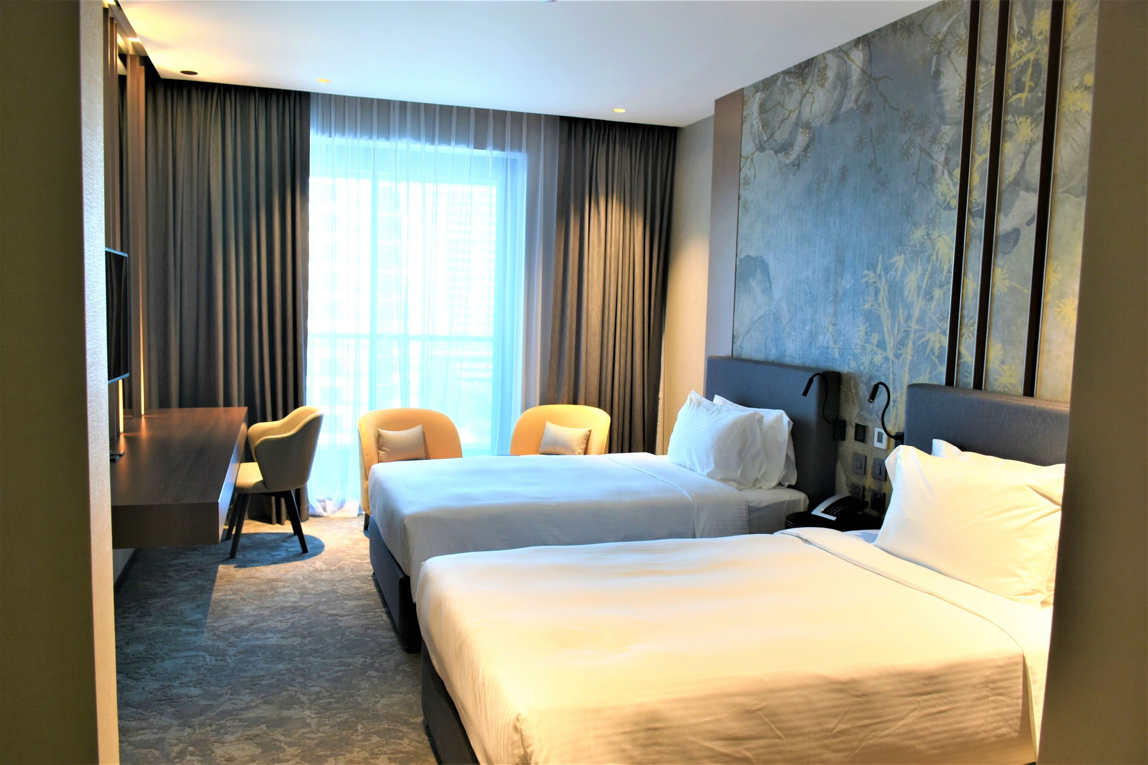 Quarto de hotel com duas camas, uma escrivaninha, uma cadeira e uma janela grande com cortinas.