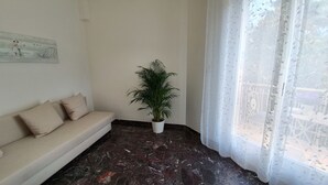 Uma sala de estar com um sofá bege, uma planta em vaso e uma janela grande com cortinas translúcidas.