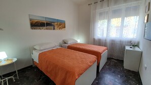 Quarto de hotel com duas camas, uma pequena escrivaninha, uma luminária e uma janela com cortinas finas.