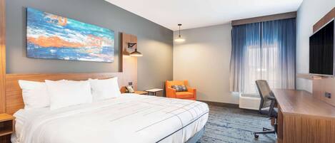 Quarto de hotel com uma cama grande, uma escrivaninha, uma cadeira, uma televisão, um quadro na parede e uma janela com cortinas.