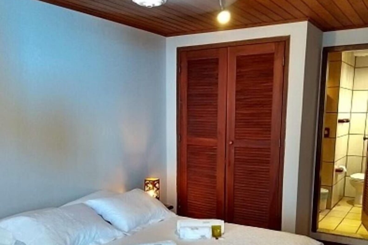 Quarto com cama, armário de madeira e banheiro visível por uma porta aberta.