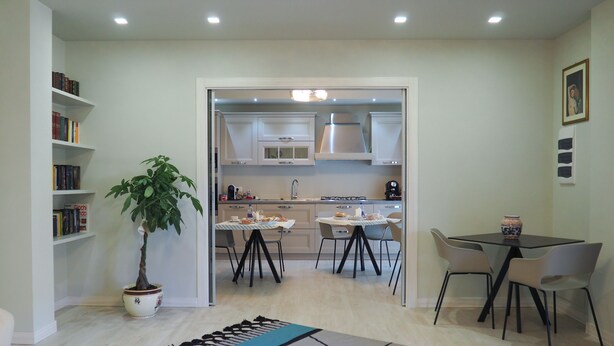 Uma sala de estar moderna com sofá, mesa de centro e área de jantar com cadeiras e mesa. Há uma estante com livros e uma planta em vaso.