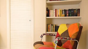 Uma poltrona colorida com estrutura metálica, posicionada ao lado de uma estante cheia de livros.