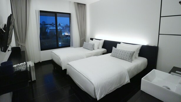 Quarto de hotel com duas camas, uma janela grande com cortinas, uma televisão de tela plana e uma mesa de cabeceira branca.