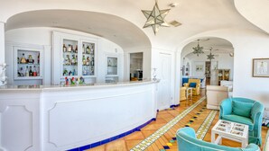 Uma área de bar com balcão branco, prateleiras com garrafas e piso estampado. Há cadeiras cor turquesa e uma mesa branca com um copo.