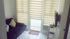 Uma sala de estar com um sofá escuro, uma televisão na parede e uma janela com persianas.