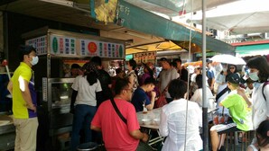 Mercado ao ar livre movimentado, com barraquinhas de comida e pessoas em fila.
