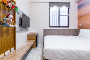 Quarto de hotel com cama, televisão fixada na parede, janela com cortinas e prateleira com diversos objetos.