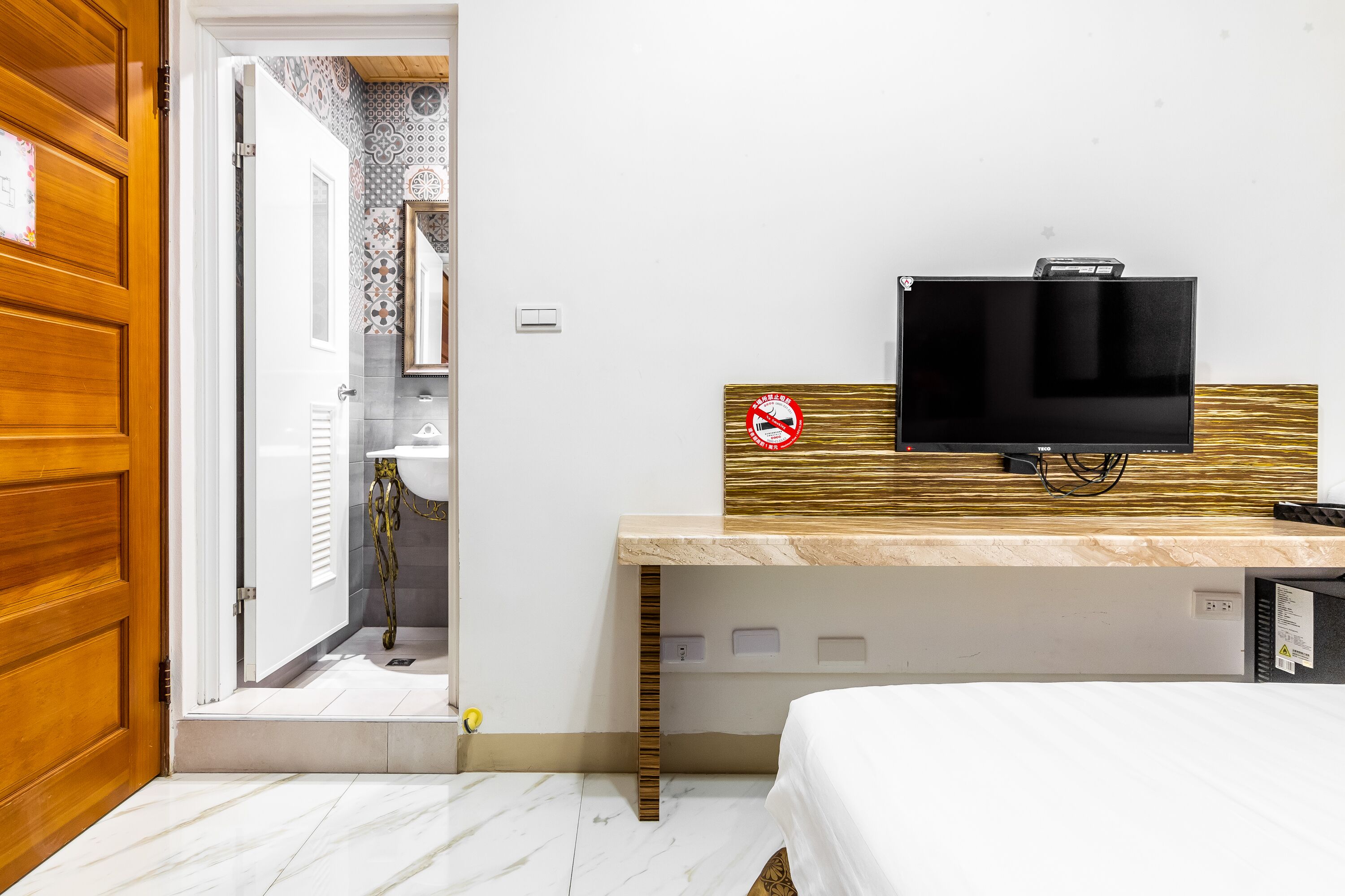 Quarto de hotel com cama, televisão presa a um painel de madeira, banheiro com chuveiro e porta que dá para outro cômodo.