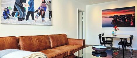 Uma sala de estar moderna com um sofá de couro marrom, uma mesa de centro de vidro e uma área de jantar com mesa e cadeiras. Há duas pinturas grandes nas paredes.