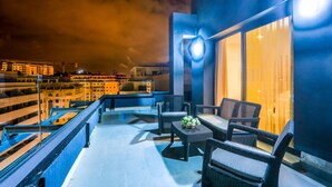 Varanda com móveis de vime e uma mesa, com vista para o horizonte urbano à noite.