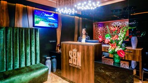 Balcão de recepção de hotel com uma televisão exibindo uma cena tropical, um candelabro moderno e um arranjo floral.