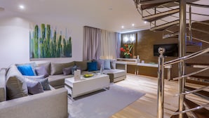 Uma sala de estar moderna com um sofá, mesa de centro e televisão. Escada com corrimão de metal que leva a um andar superior.