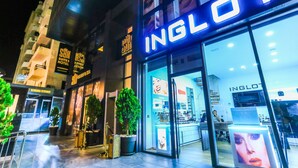 Entrada de hotel com um destaque para a placa 'INGLOT', fachada de vidro e interior visível, incluindo um balcão de recepção e vitrines.