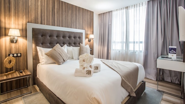 Quarto de hotel moderno com uma cama grande, paredes de painel de madeira e vista da cidade através de amplas janelas.