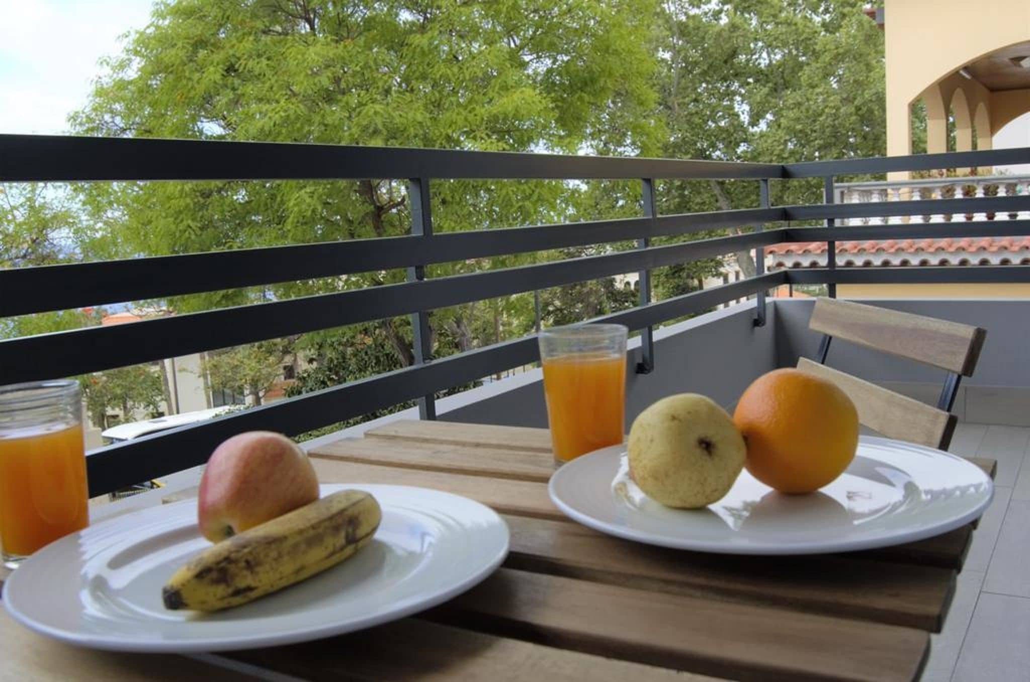 Uma varanda com uma mesa posta para o café da manhã, com pratos contendo uma banana, uma maçã e uma laranja, e copos de suco de laranja.