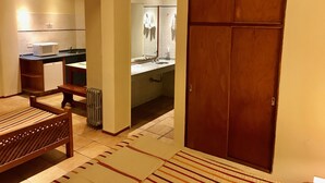 Quarto de hotel com um banco de madeira, um armário de madeira, área de cozinha com micro-ondas e banheiro com espelho e pia.