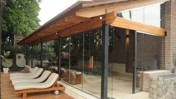 Uma área de estar externa moderna com pérgola de madeira, paredes de vidro e lareira de pedra.
