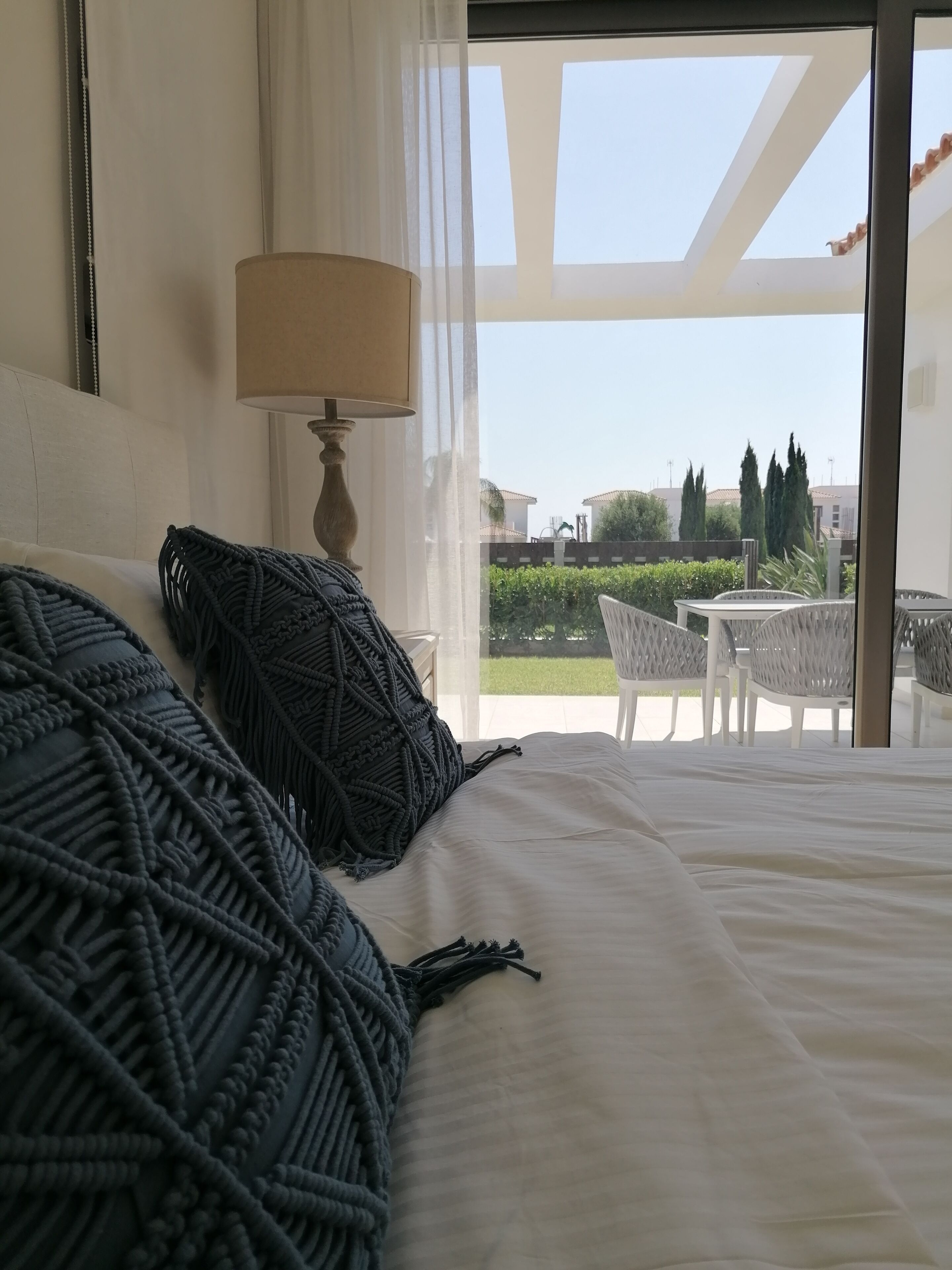 Quarto com cama, abajur e vista para um pátio externo com cadeiras e mesa.