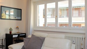 Quarto com sofá branco, escrivaninha com cadeira, quadro pendurado na parede e janela com vista para edifícios.