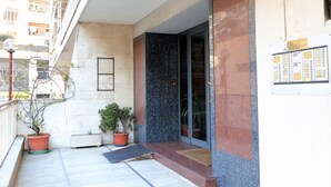 Entrada de um edifício com chão de azulejos, plantas em vasos e porta com painéis de vidro.