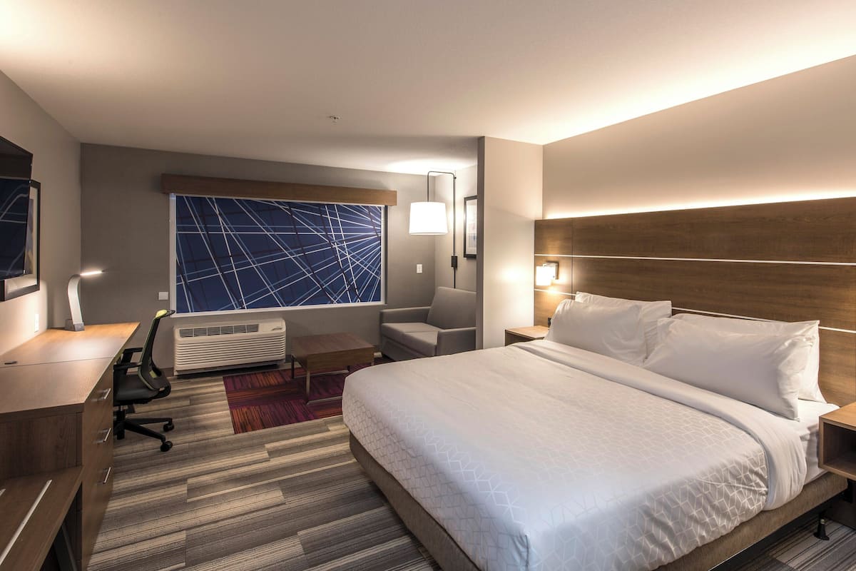 Quarto de hotel com uma cama grande, uma escrivaninha, uma cadeira, uma luminária de mesa, uma luminária de parede e um ar condicionado, além de uma grande janela com padrão geométrico.