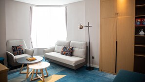 Uma sala de estar moderna com um sofá, poltrona, mesa de centro e um abajur. Há uma janela com cortinas, uma estante de livros e uma mesinha com um controle remoto.