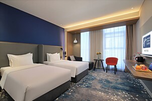 Quarto de hotel com duas camas, uma escrivaninha, uma cadeira, uma mesinha com um vaso e uma televisão na parede.