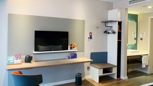 Quarto de hotel moderno com televisão de tela plana, uma pequena escrivaninha com cadeira, um banco, uma lixeira e banheiro com pia e espelho.